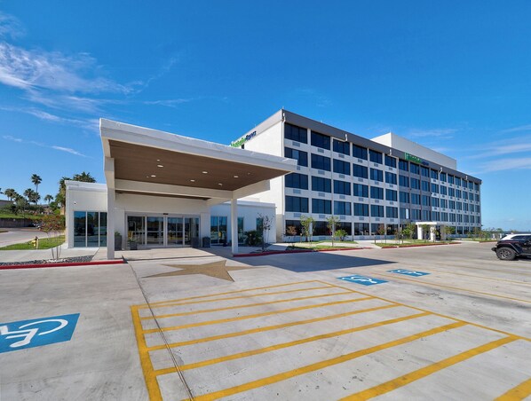 Um edifício moderno com entrada coberta, vagas de estacionamento demarcadas e um céu azul límpido.