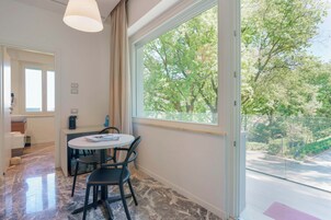 Um quarto com uma grande janela, uma mesa redonda com cadeiras, uma pequena escrivaninha e uma cadeira.