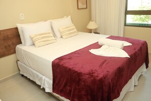 Uma cama bem arrumada com colcha branca e vinho, travesseiros brancos e uma toalha branca dobrada ao lado.