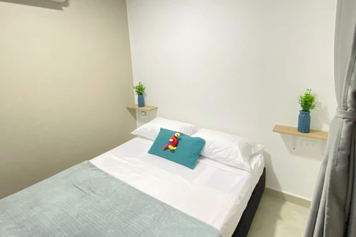Uma cama bem arrumada com um travesseiro azul-turquesa e um cobertor azul claro, duas pequenas mesas de cabeceira com plantas em vasos, e uma cortina cinza na janela.