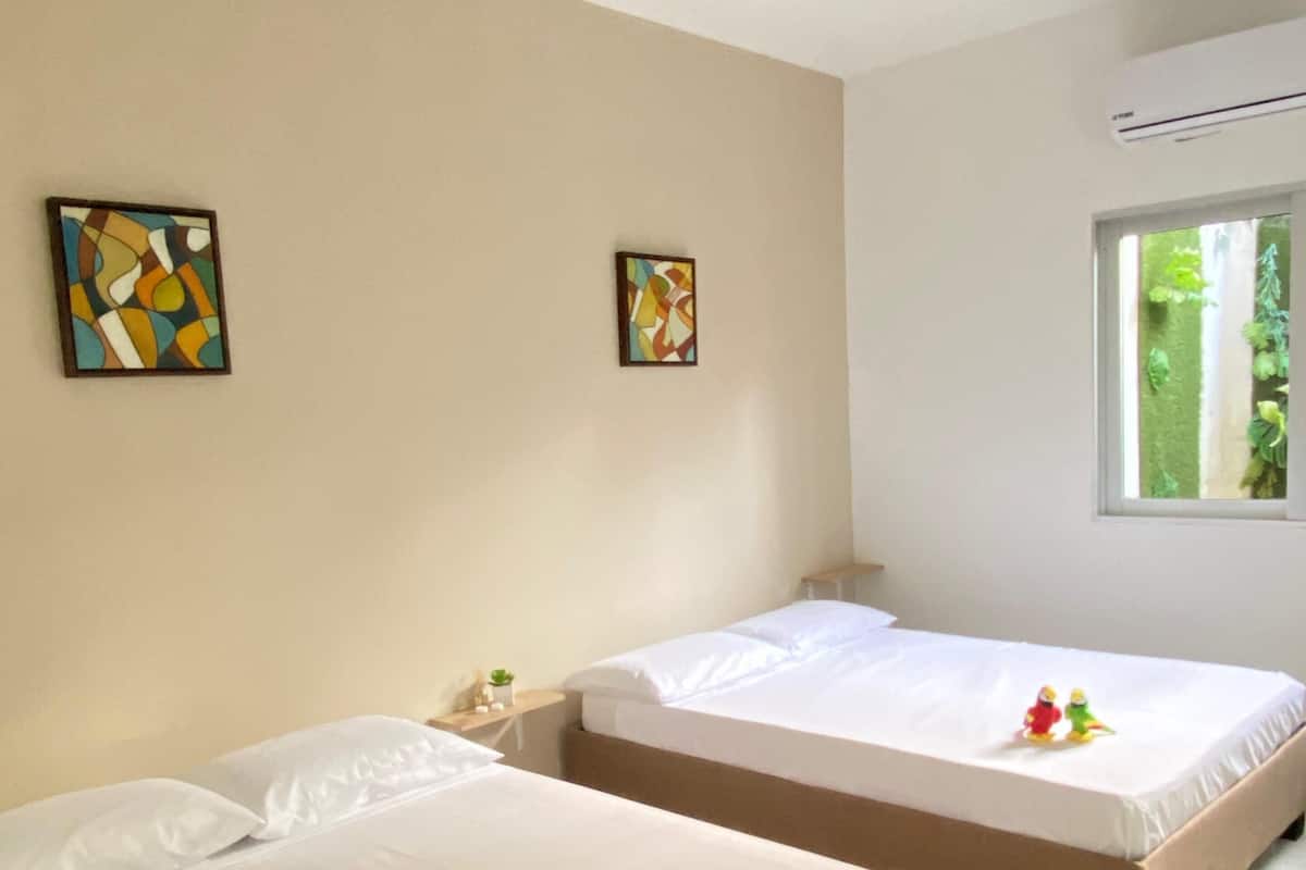 Quarto de hotel com duas camas, um ventilador de teto, obras de arte nas paredes e janela com vista para o verde.