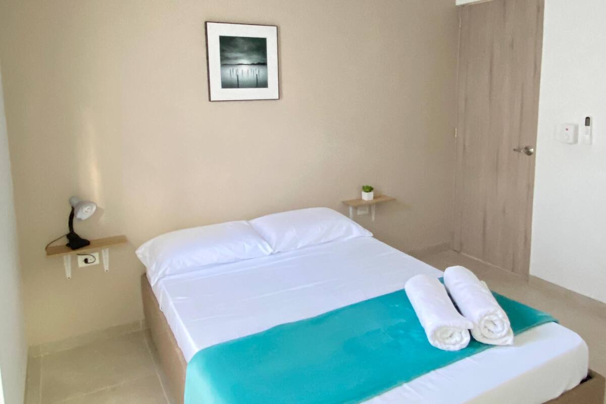Cama bem arrumada com manta verde-água e lençóis brancos, uma pequena mesa de cabeceira com luminária, uma moldura na parede e um ventilador de teto.
