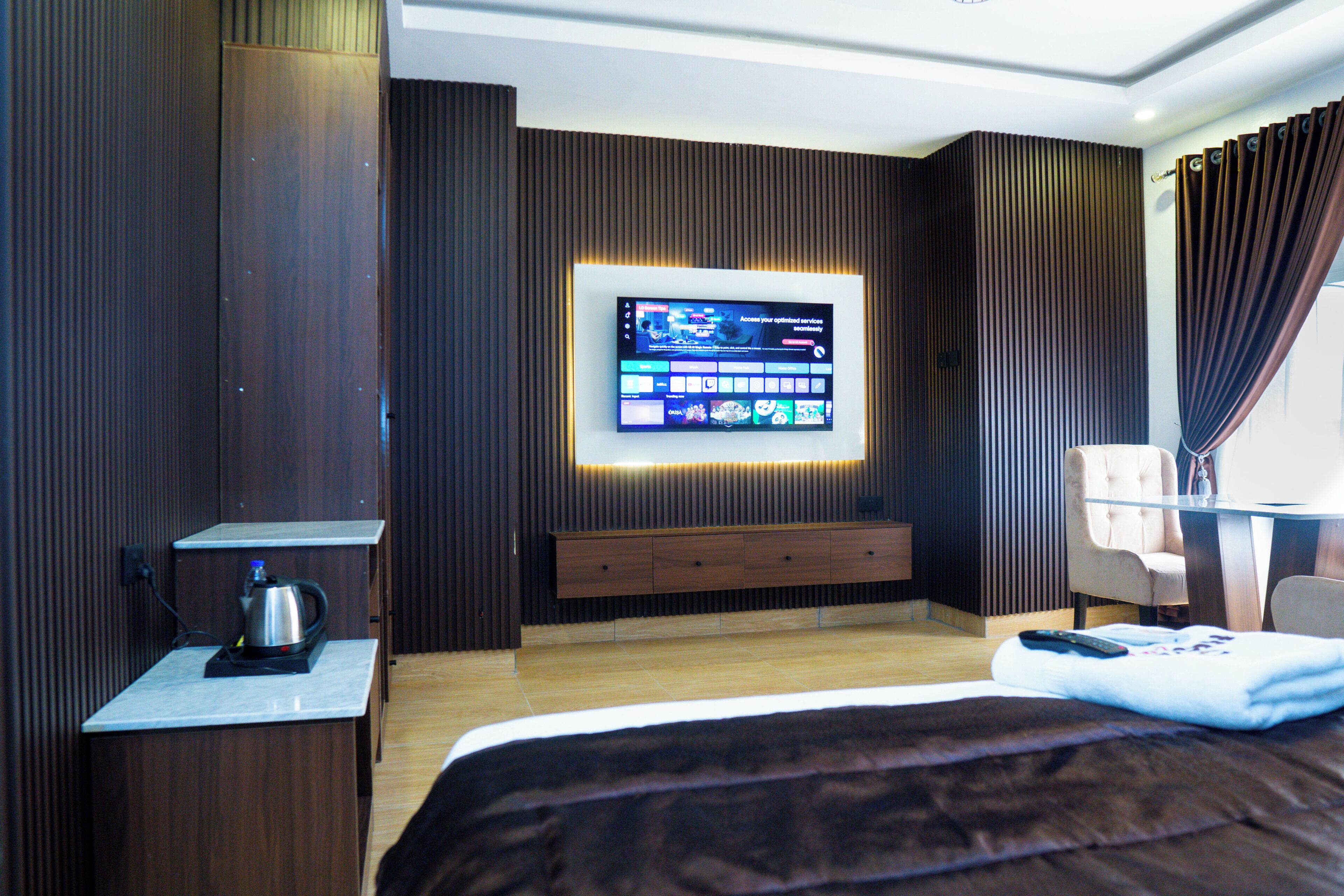 Quarto de hotel com cama, televisão na parede, secretária com chaleira e uma cadeira.