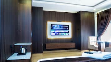 Quarto de hotel com cama, televisão na parede, secretária com chaleira e uma cadeira.