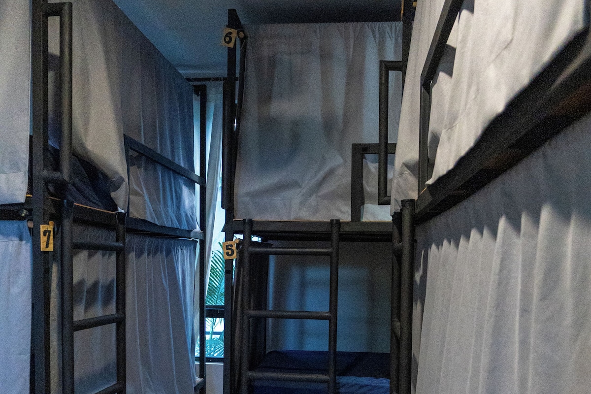 Um corredor estreito com beliches em ambos os lados, um ventilador no teto e uma janela no fim.