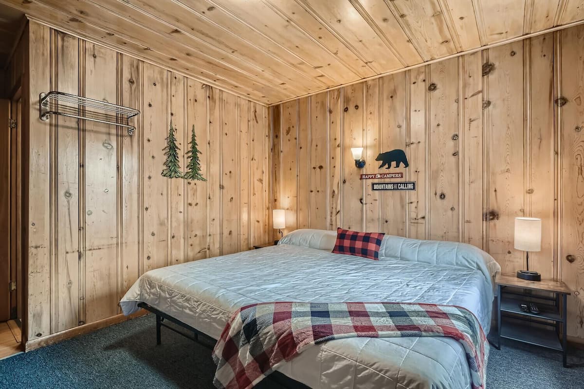 Quarto com painéis de madeira, com uma cama, um porta-toalhas de metal, uma luminária de parede e um letreiro que diz "HAPPY CAMPERS MOUNTAINS ARE CALLING".