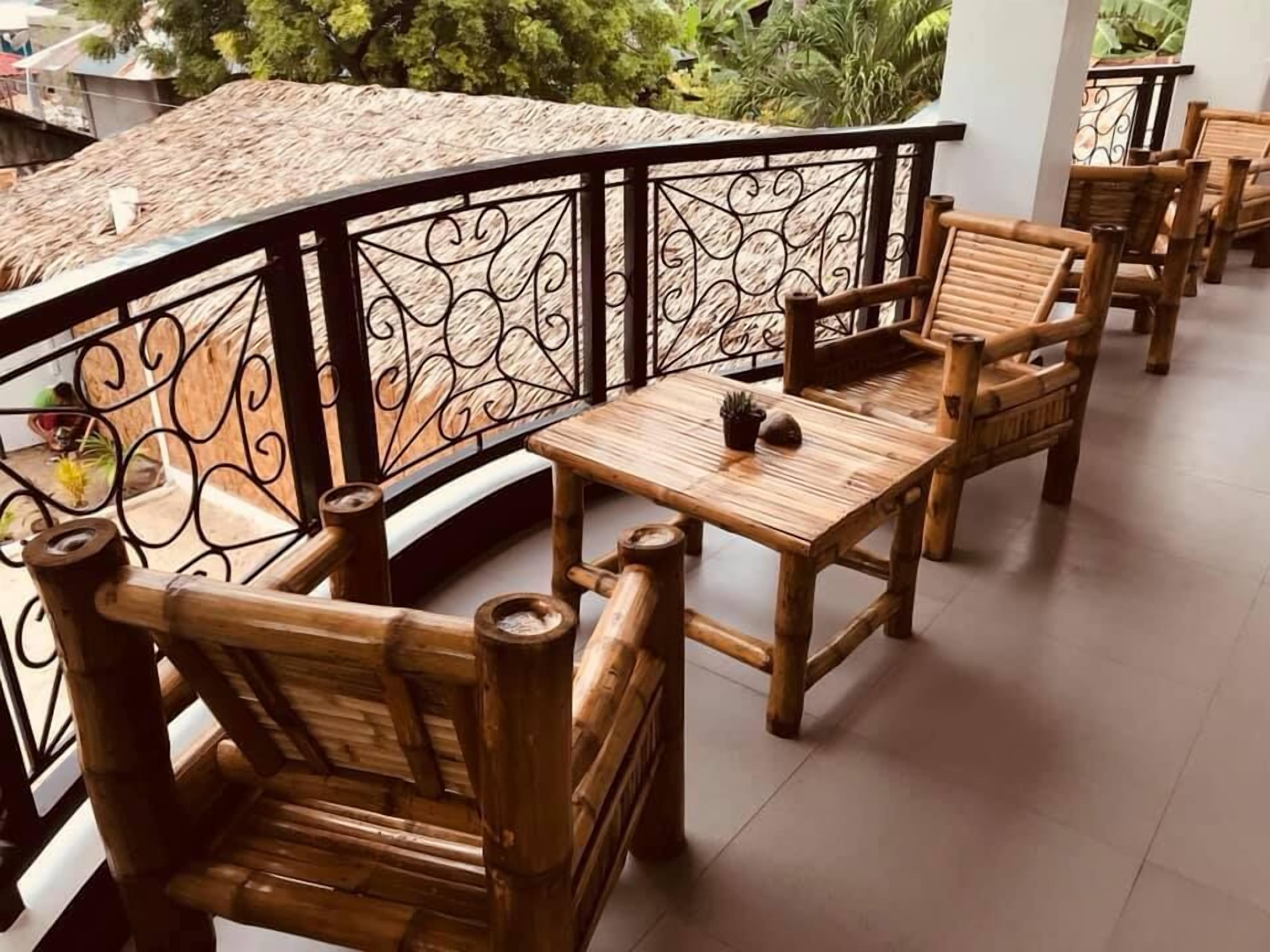 Um deck de madeira com guarda-corpo de metal, rodeado por cadeiras e mesas de bambu, e vegetação ao fundo.