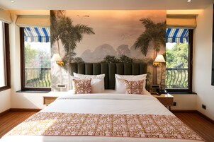 Quarto de hotel com uma cama grande, duas mesas de cabeceira, uma luminária na parede e vista para palmeiras e montanhas pelas janelas.