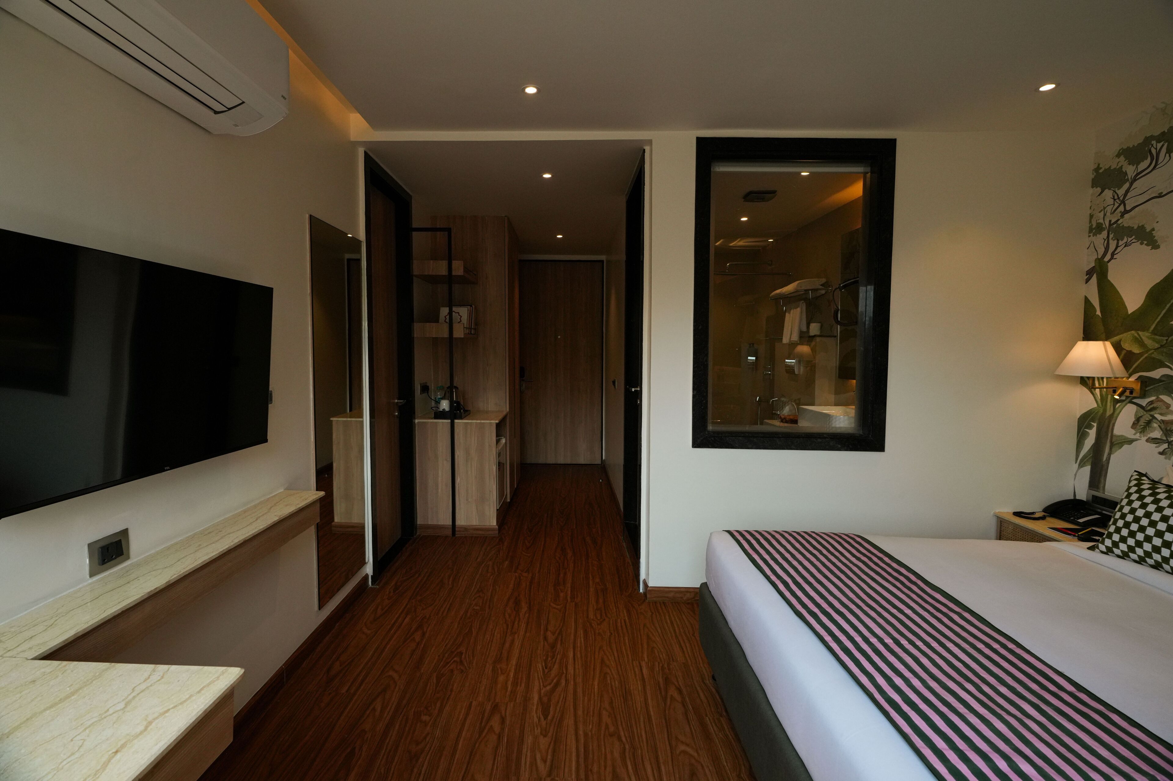 Quarto de hotel com uma televisão de tela plana grande, uma cama com roupa de cama listrada, uma mesa de cabeceira com abajur e um espelho na parede.