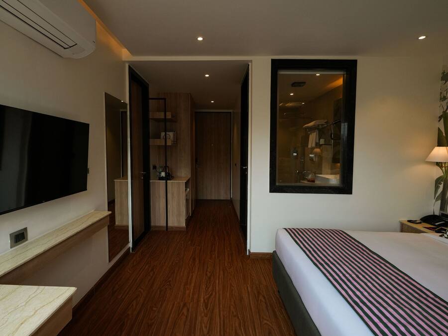 Quarto de hotel com uma televisão de tela plana grande, uma cama com roupa de cama listrada, uma mesa de cabeceira com abajur e um espelho na parede.