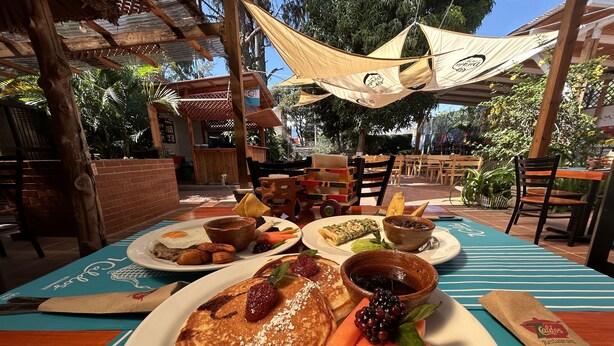 Uma mesa de jantar com diversas opções de pequeno-almoço, incluindo panquecas, uma taça de fruta e um prato com ovos e torradas.