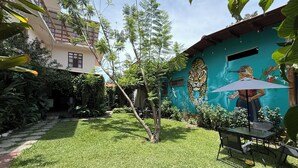 Um pátio com um mural de um leopardo e um beija-flor, rodeado por vegetação exuberante e uma pequena mesa com cadeiras.