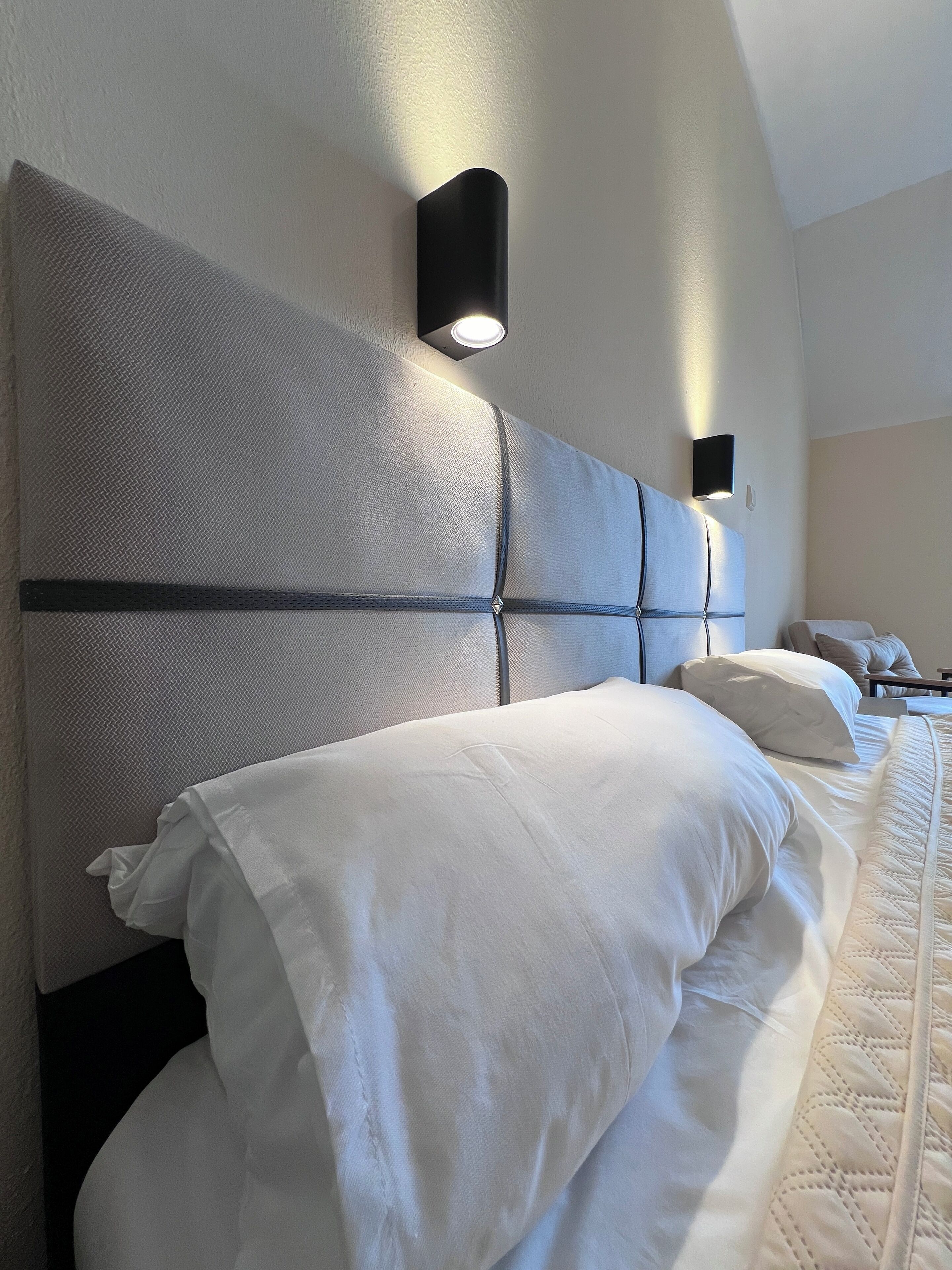 Quarto de hotel moderno com uma cama grande, cabeceira com design texturizado e duas luminárias fixadas na parede.
