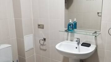 Uma casa de banho com sanita, lavatório sobreposta e uma prateleira de vidro com produtos de higiene pessoal, além de um espelho.
