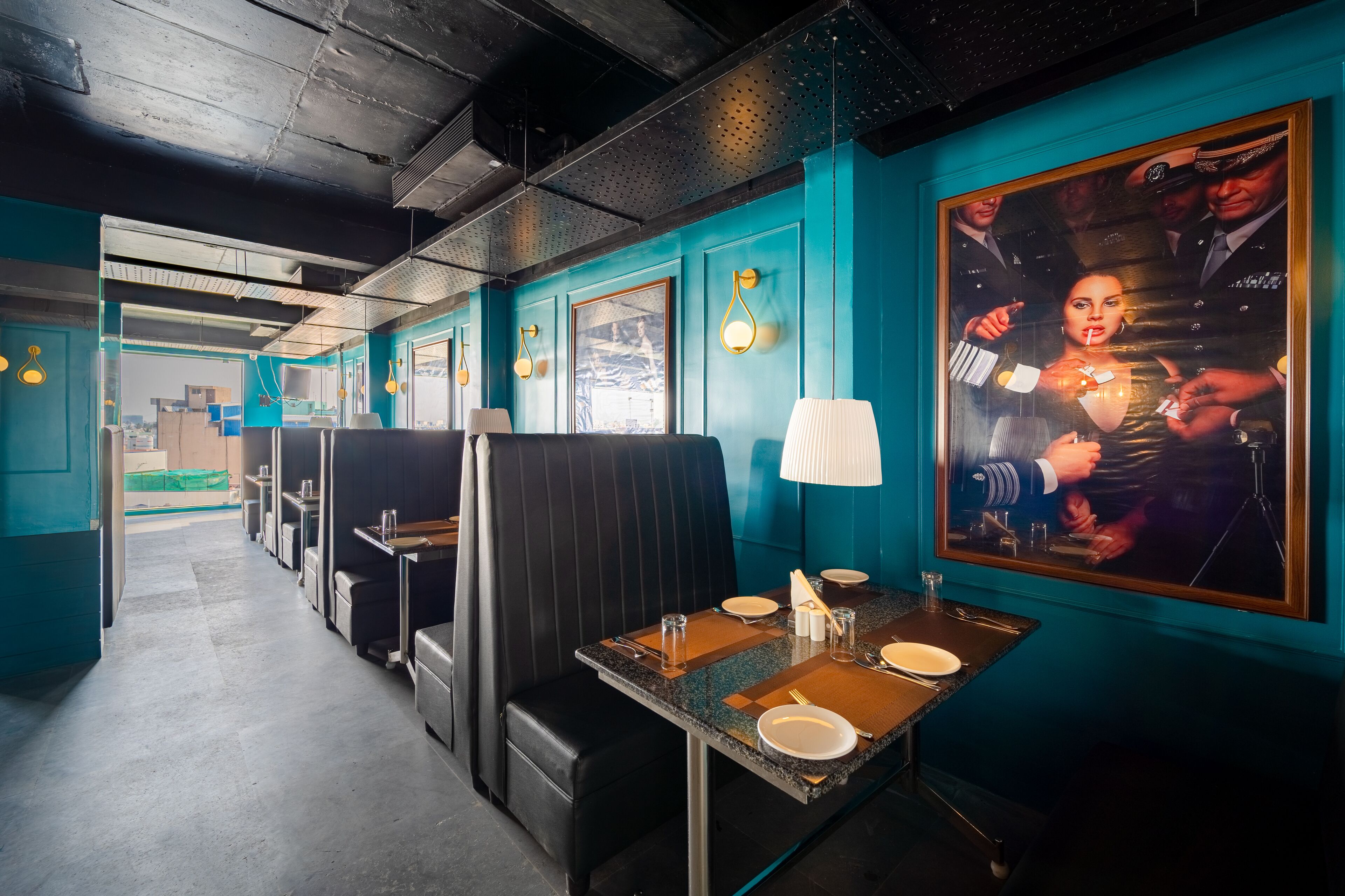 Um restaurante com paredes verde-água, cabines pretas e uma grande pintura retratando um grupo de pessoas. As mesas estão postas com pratos, copos e talheres.