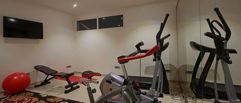 Uma academia em casa com equipamentos de ginástica, incluindo uma bicicleta ergométrica, elíptico e uma bola de exercício vermelha.