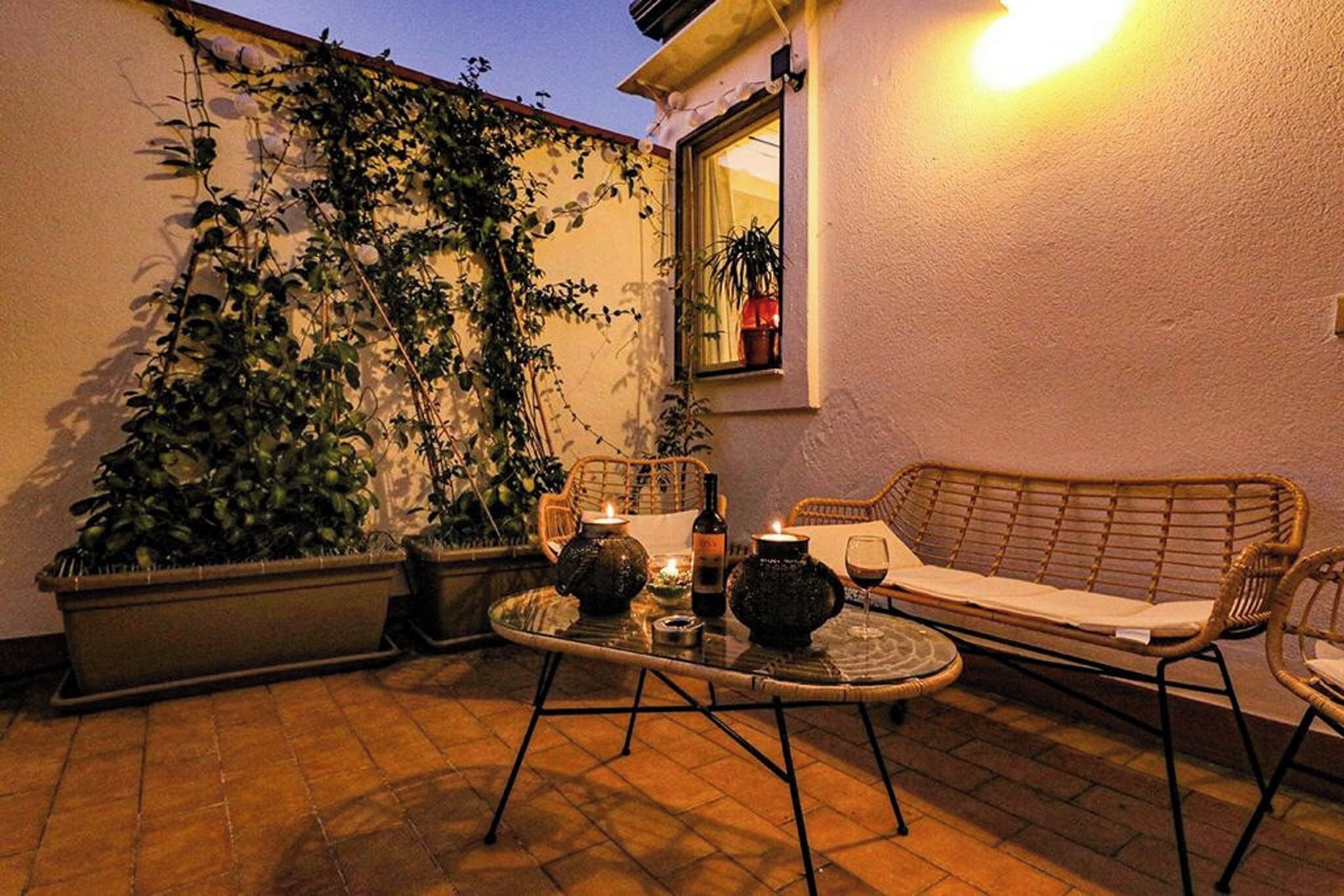 Um pátio externo com um sofá de vime, uma mesa redonda, plantas em vasos e uma janela com vista.