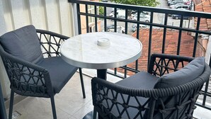 Varanda com duas cadeiras de vime e uma mesa redonda pequena, com vista para um parque de estacionamento e edifícios.