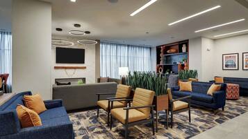 Um saguão de hotel moderno com um sofá, poltronas, uma televisão de tela plana, uma mesa de centro e uma grande planta em vaso.