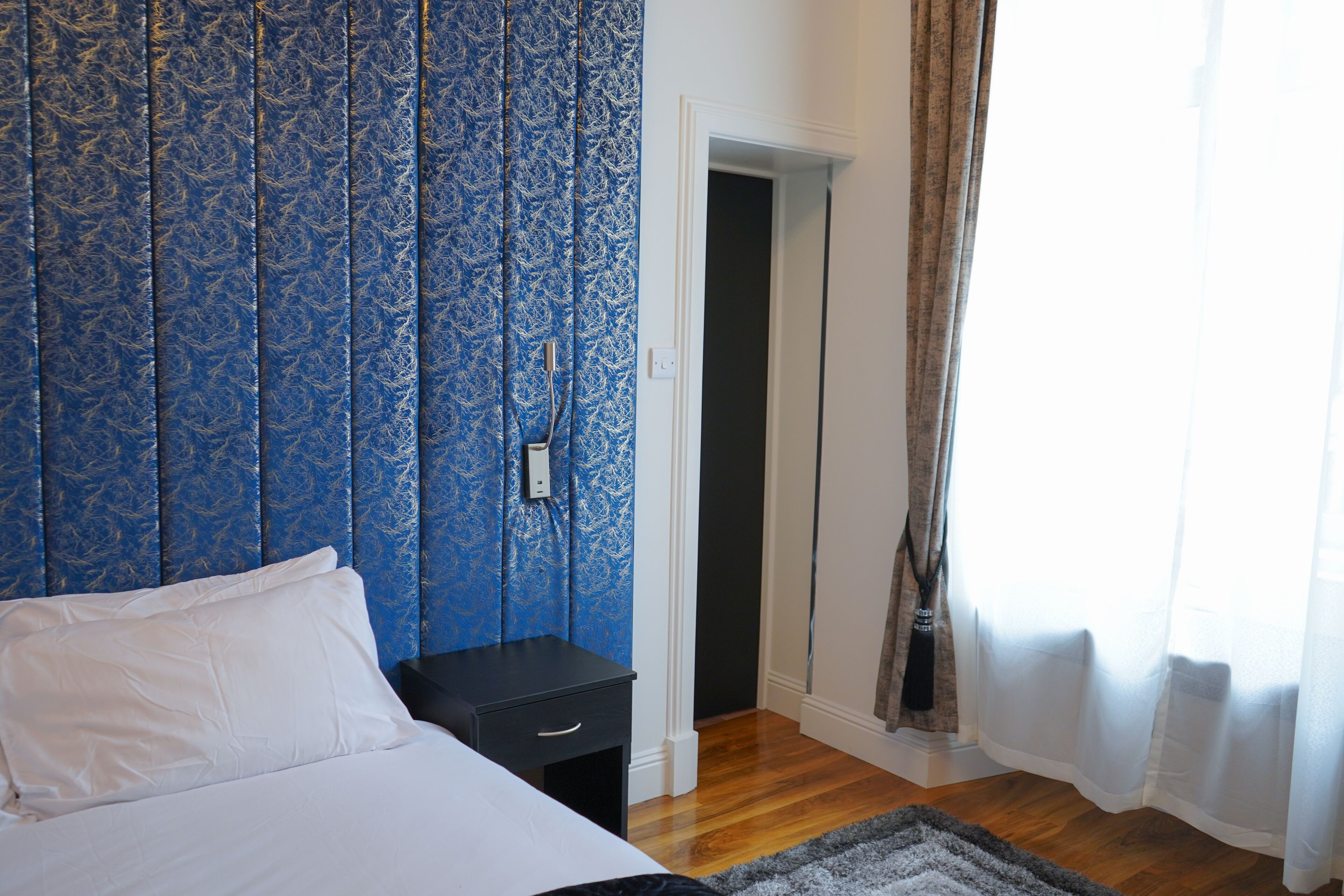 Quarto com cama, mesa de cabeceira, porta e janela com cortinas.