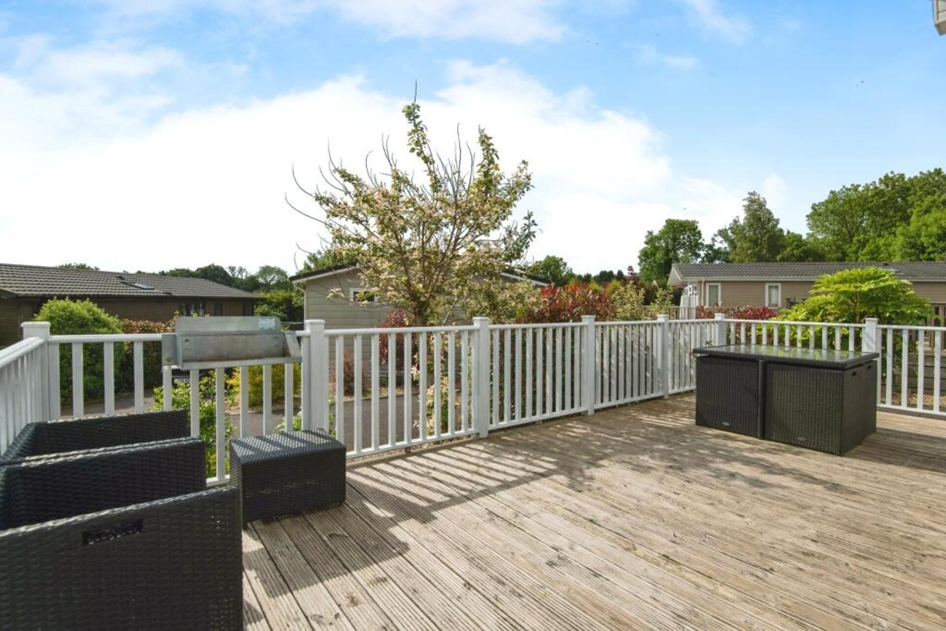 Um deck de madeira com guarda-corpos brancos, móveis de vime e vista para casas e árvores vizinhas.