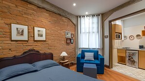 Quarto com parede de tijolos, uma cama, uma poltrona azul, uma mesinha de cabeceira com abajur e uma janela com cortinas.