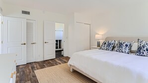 Quarto com cama, paredes brancas, soalho de madeira, luz no teto e uma porta.