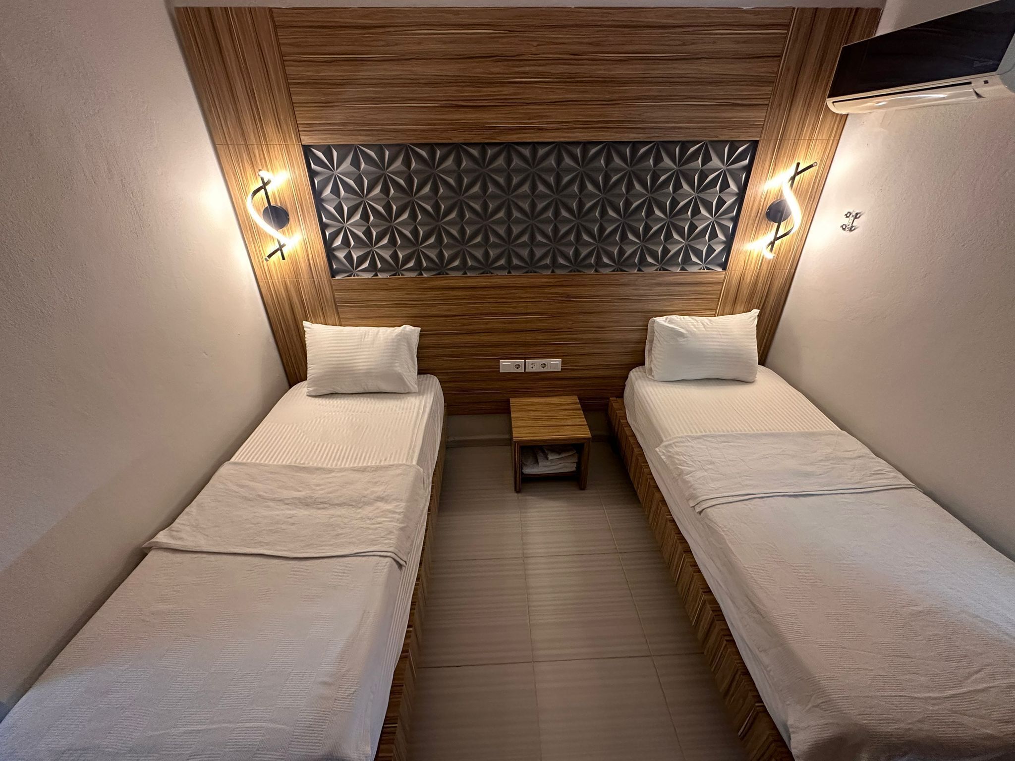Um quarto pequeno e aconchegante com duas camas de solteiro, uma cabeceira de madeira e um painel decorativo na parede.