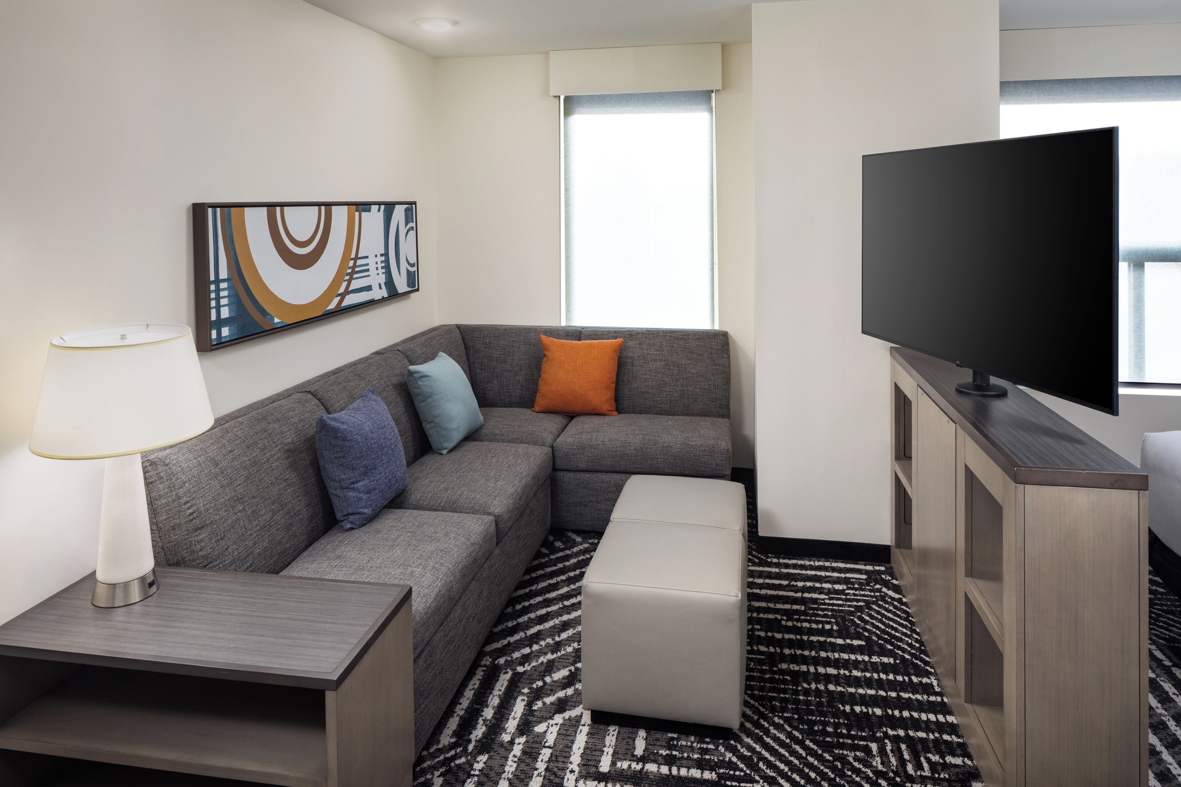 Quarto de hotel moderno com sofá modular cinza, televisão de tela plana, luminária e tapete estampado.