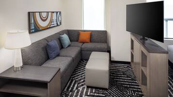 Quarto de hotel moderno com sofá modular cinza, televisão de tela plana, luminária e tapete estampado.