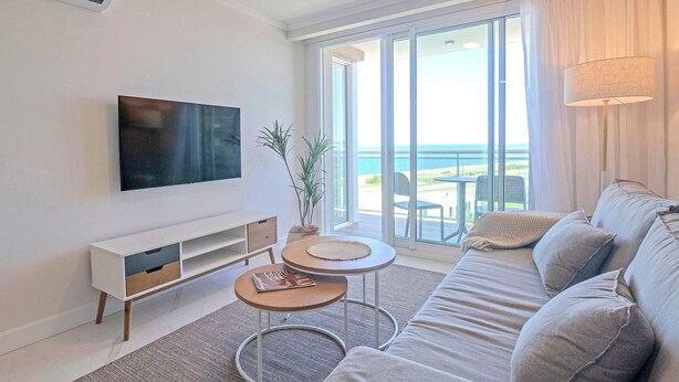 Sala de estar moderna com televisão de tela plana, sofá, duas mesinhas e uma grande porta de vidro deslizante que dá para uma varanda com vista para o mar.