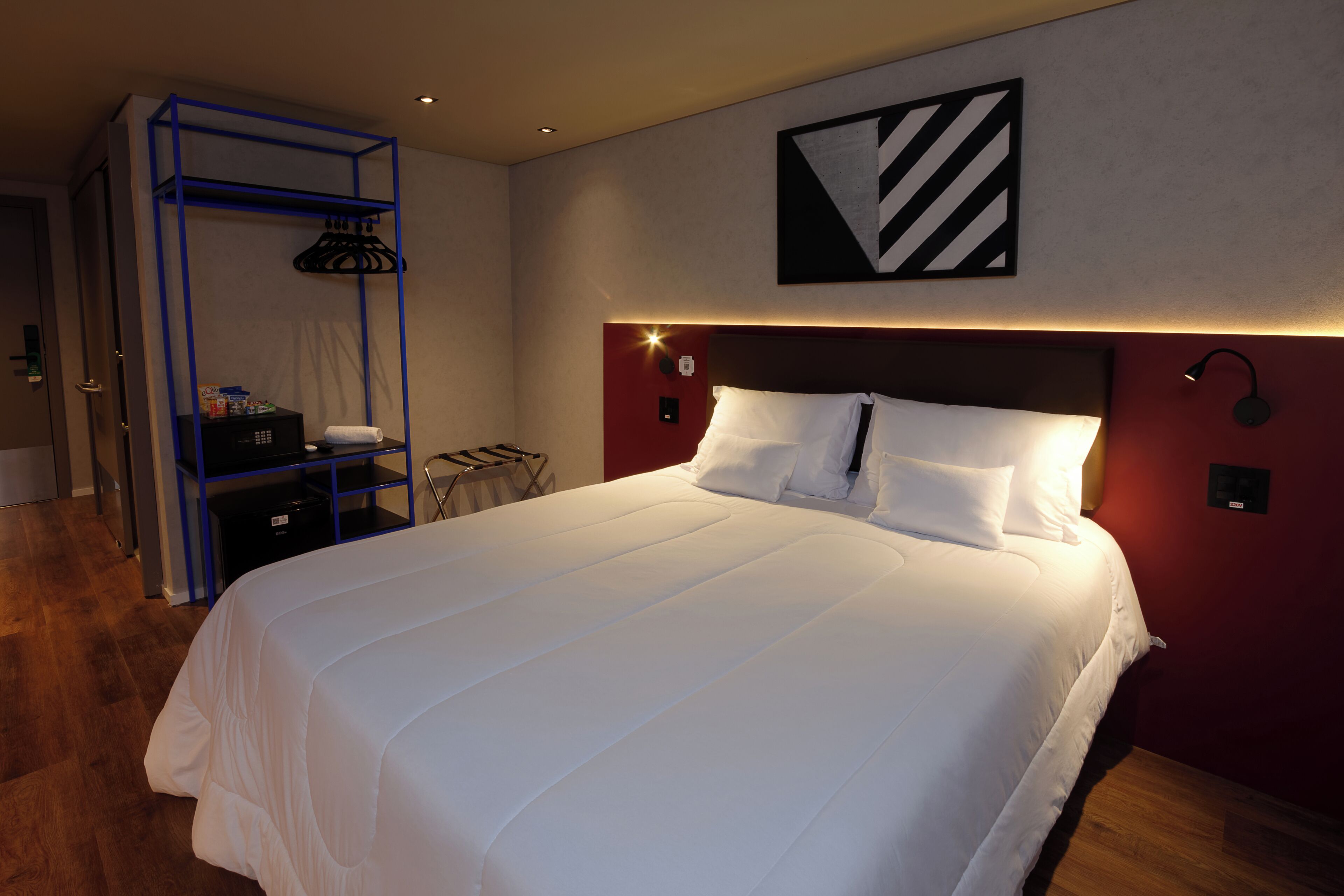 Quarto de hotel com uma cama grande, um móvel de armazenamento azul, uma luminária fixada na parede e uma obra de arte geométrica.