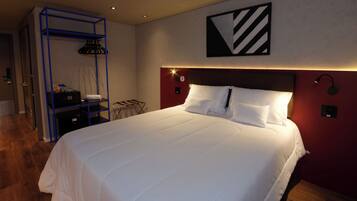 Quarto de hotel com uma cama grande, um móvel de armazenamento azul, uma luminária fixada na parede e uma obra de arte geométrica.