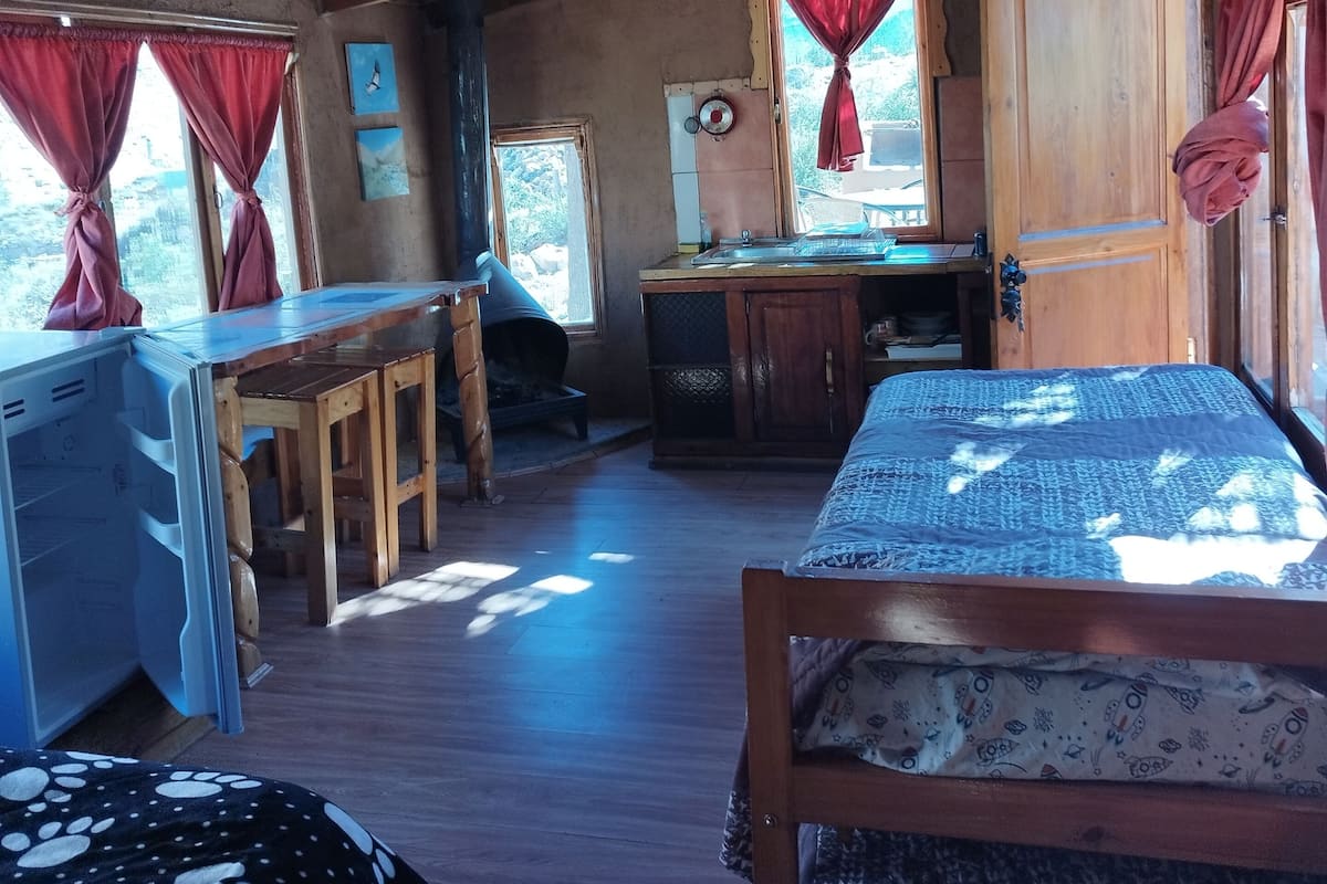 Interior rústico de cabana com cama de madeira, mesa de jantar, pequena cozinha e lareira.