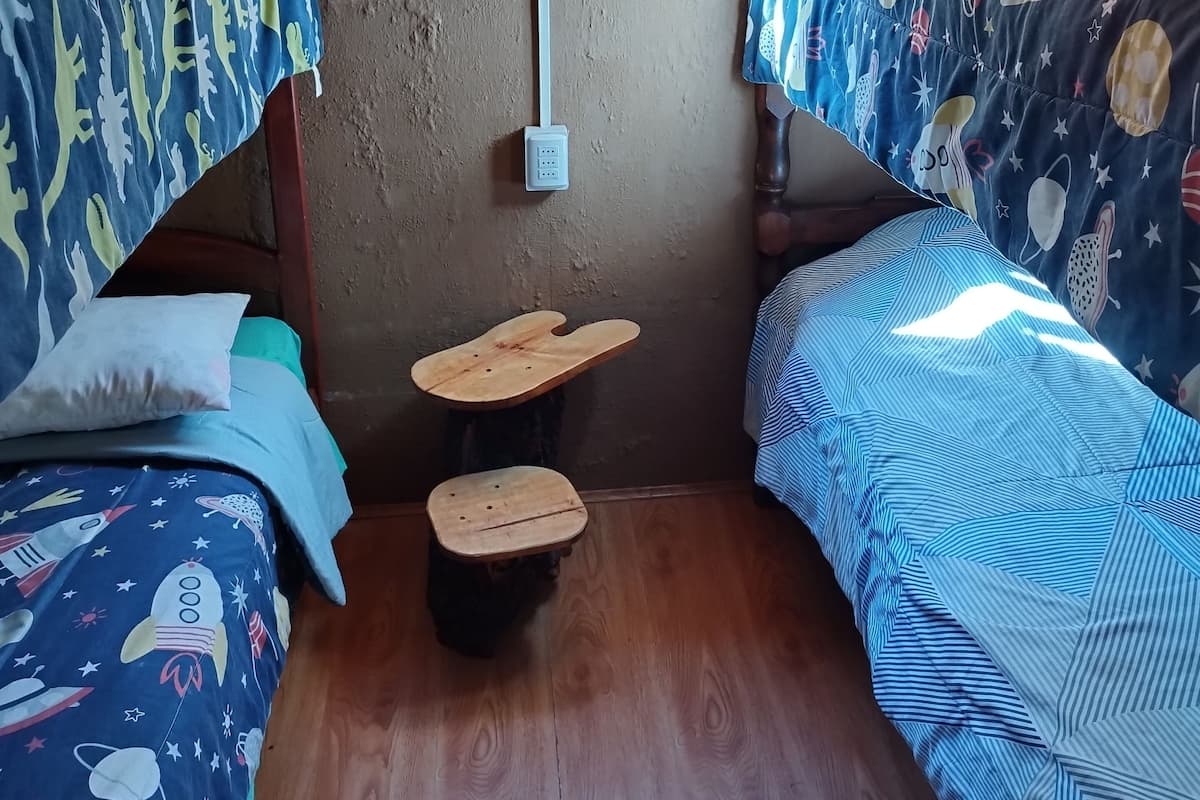 Um quarto pequeno e acolhedor com duas camas, uma janela e um banquinho de madeira.
