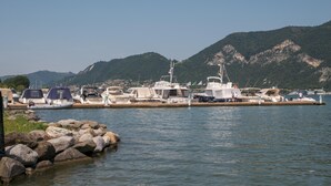 Uma marina com barcos atracados, costa rochosa e montanhas ao fundo.