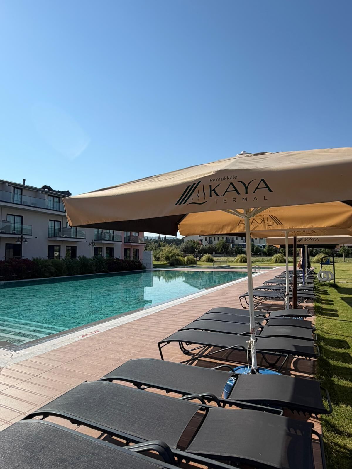 Uma área à beira da piscina com espreguiçadeiras e guarda-sóis, piscina translúcida e um edifício ao fundo.