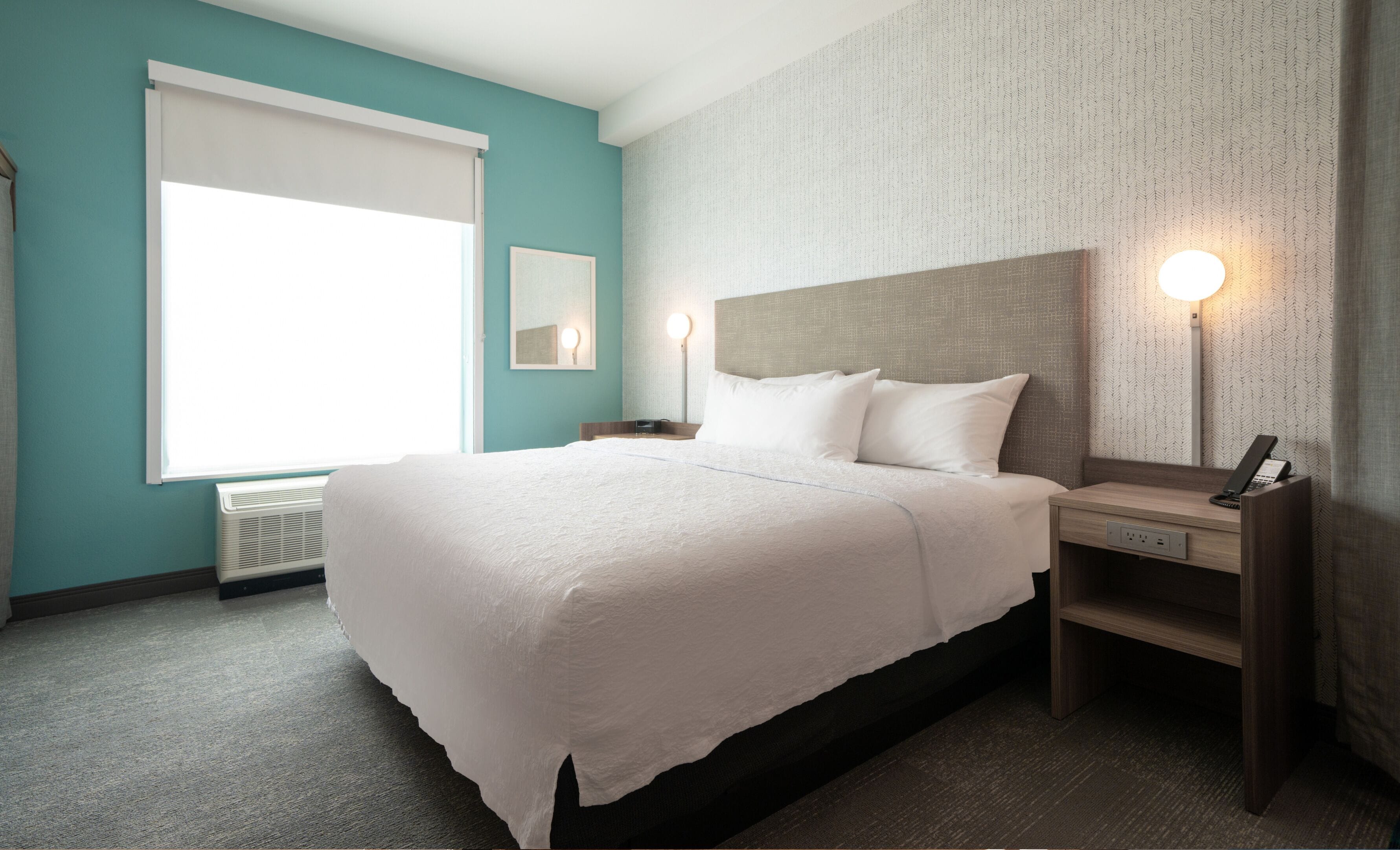 Quarto de hotel com cama grande, mesa de cabeceira com luminária, espelho fixado na parede e janela com persianas.