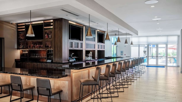 Uma área de bar moderna com um balcão comprido, vários bancos altos e luminárias pendentes.
