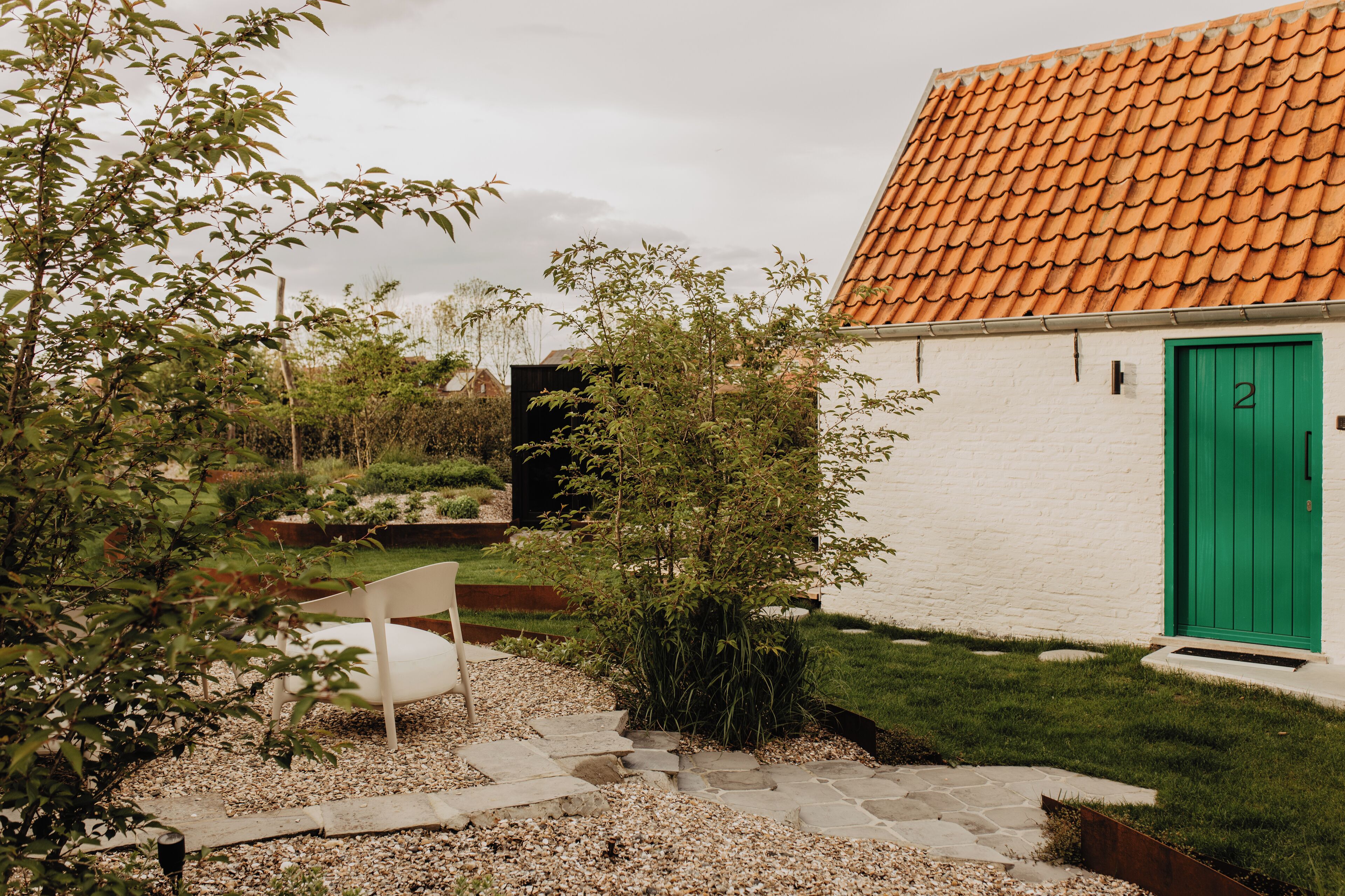 Um edifício branco com porta verde, telhado de telhas e jardim com cadeira branca e caminho de brita.