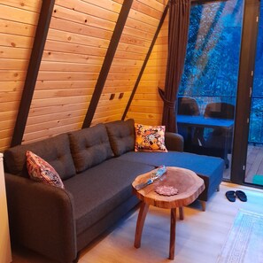 Quarto acolhedor com um sofá marrom escuro, duas almofadas estampadas, uma pequena mesa de madeira com uma faca e um prato decorativo, e uma porta de vidro que dá para o exterior.