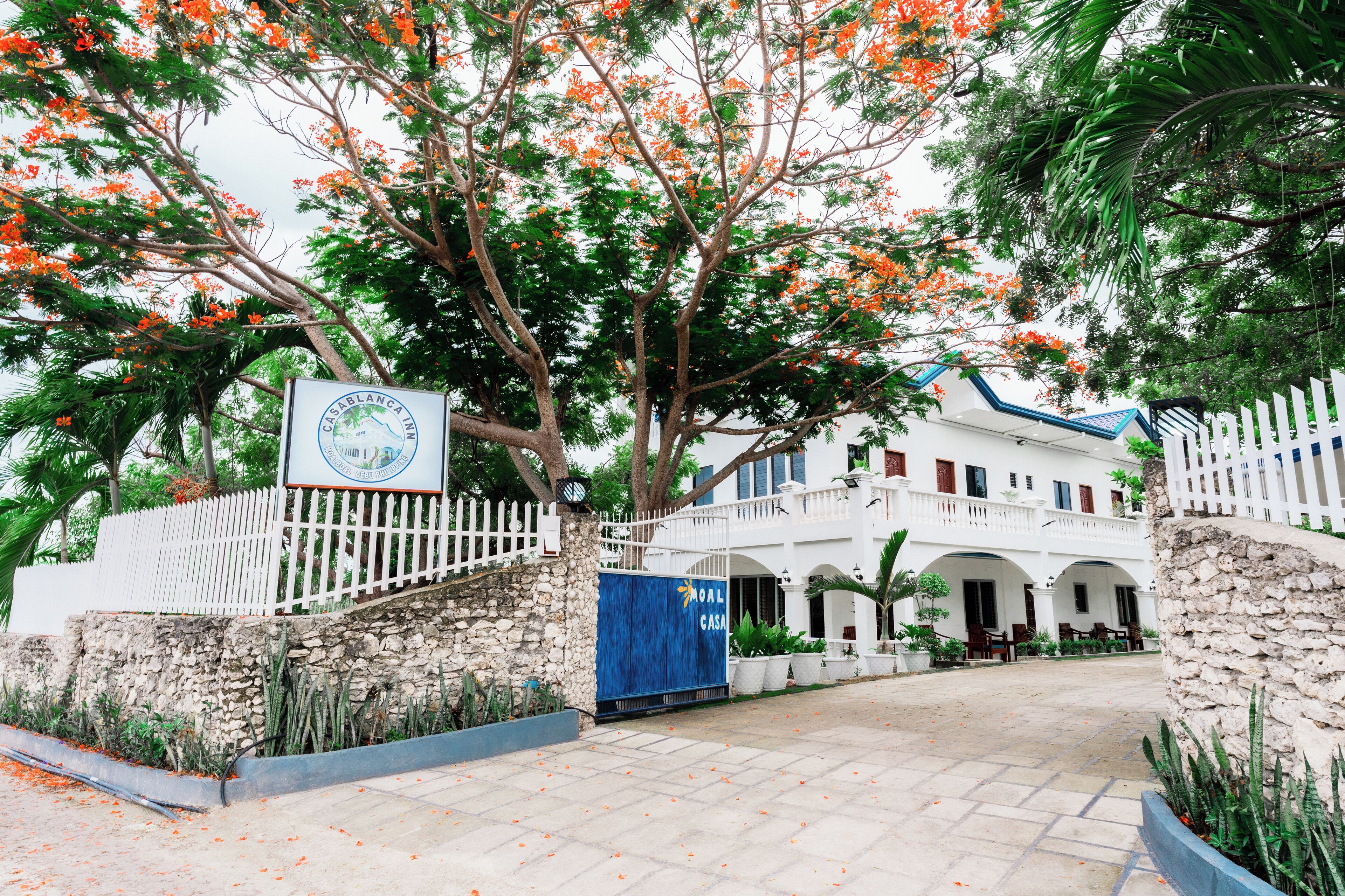Entrada de hotel com parede de pedra, cerca branca e portão azul.