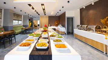 Disposição de buffet com diversas estações de comida, área de estar com mesas e cadeiras, e arranjo decorativo de plantas.