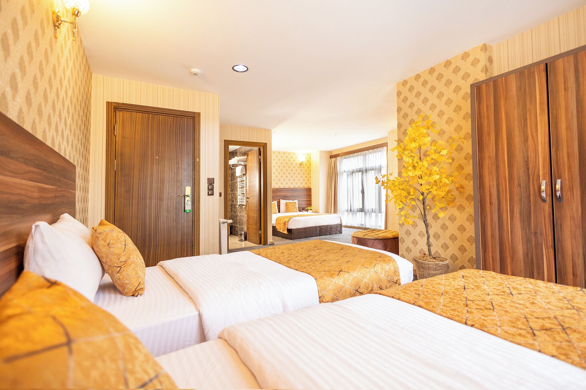 Quarto de hotel com três camas, guarda-roupas de madeira, um pequeno espaço de estar e uma planta em vaso.