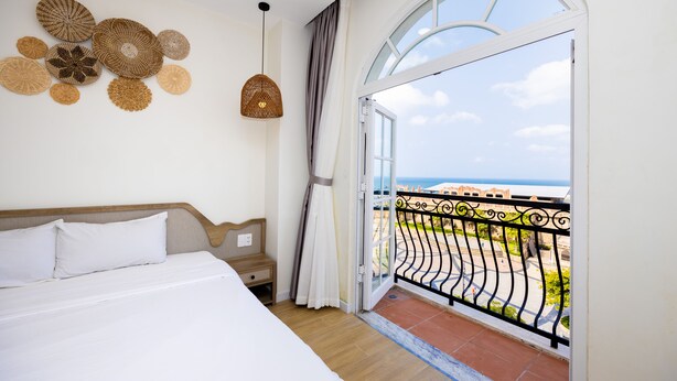 Quarto de hotel com cama, luminária fixada na parede, vista para o mar através de uma porta de varanda aberta e um corrimão.