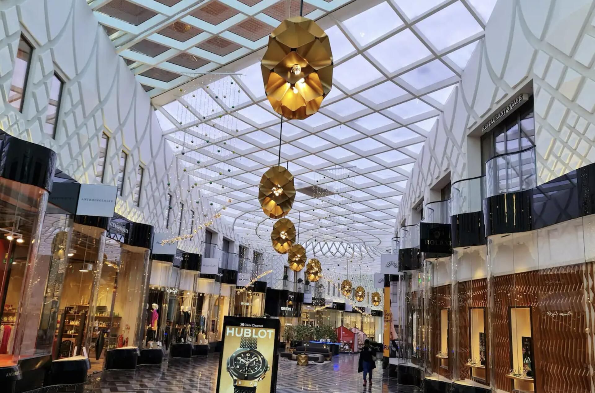 Ampla área comercial interna com teto alto, painéis de vidro e luminárias decorativas pendentes.