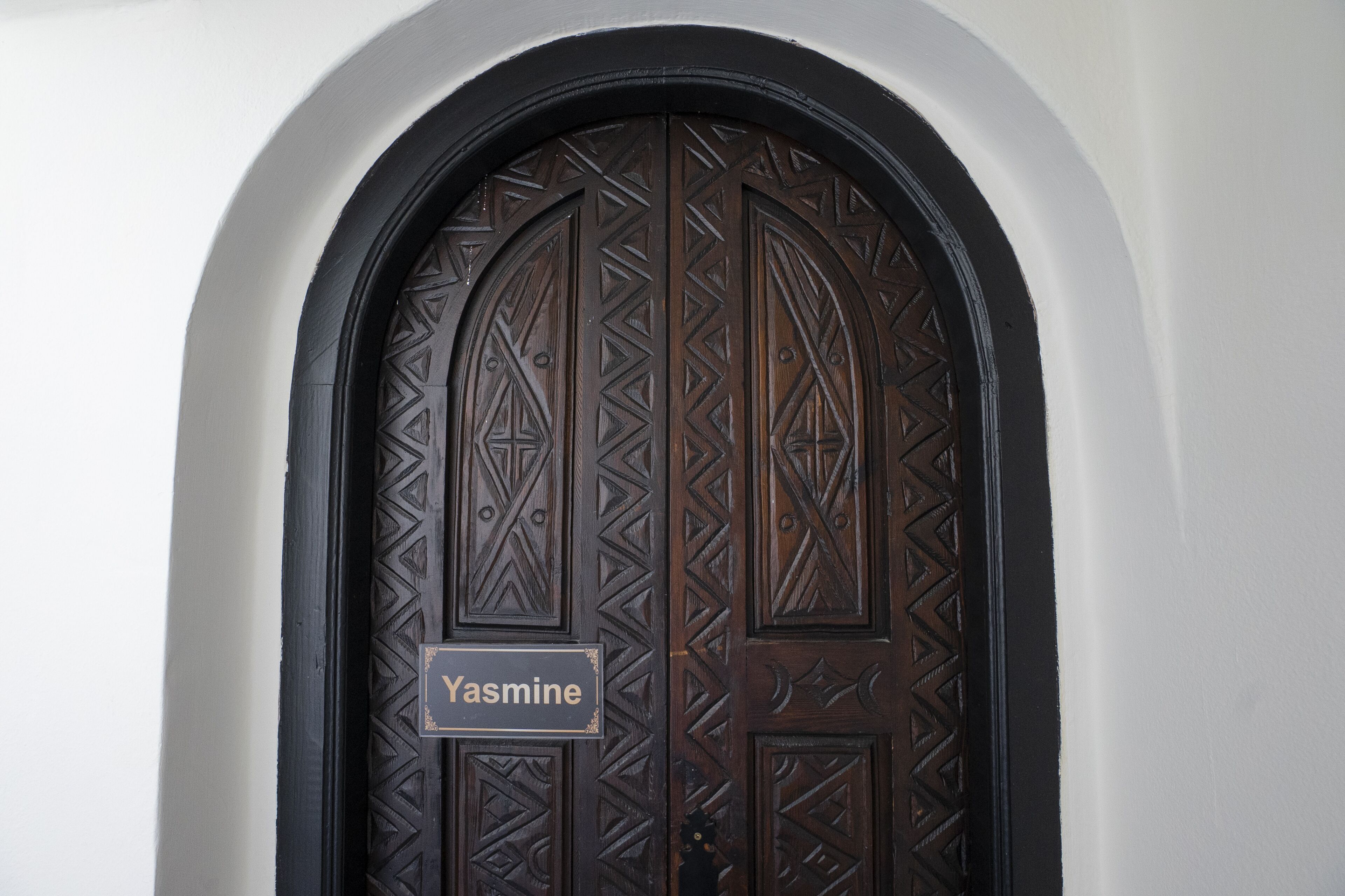 Uma porta de madeira escura com uma placa que diz "Yasmine" e uma parede branca em arco.