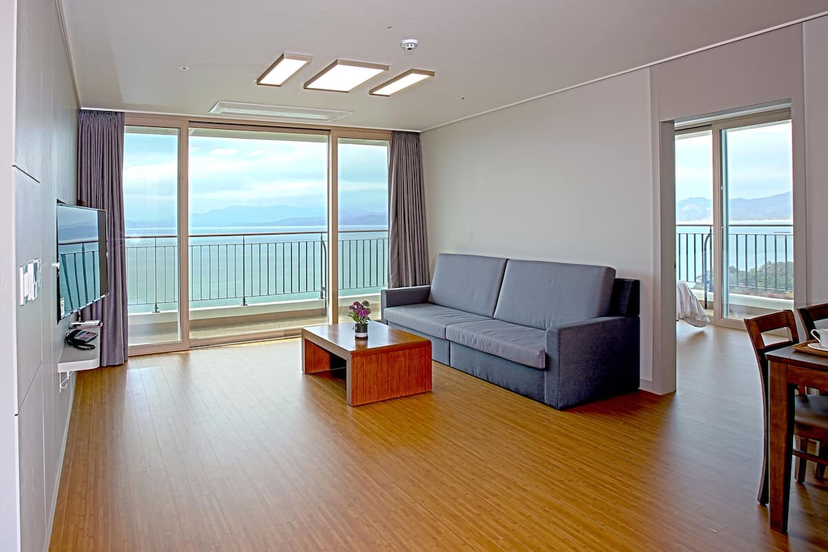 Uma sala de estar moderna com um sofá cinzento, mesa de centro de madeira e uma mesa de jantar com cadeiras. Grandes janelas proporcionam vista para o mar e para as montanhas.