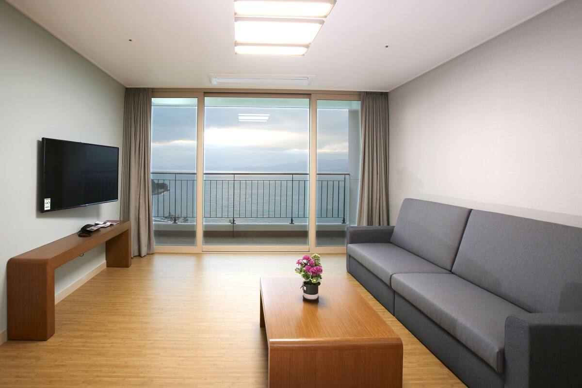 Uma sala de estar moderna com televisão de tela plana, um sofá cinza, uma mesa de centro de madeira com um vaso de flores, um banco de madeira e uma varanda com guarda-corrimão.