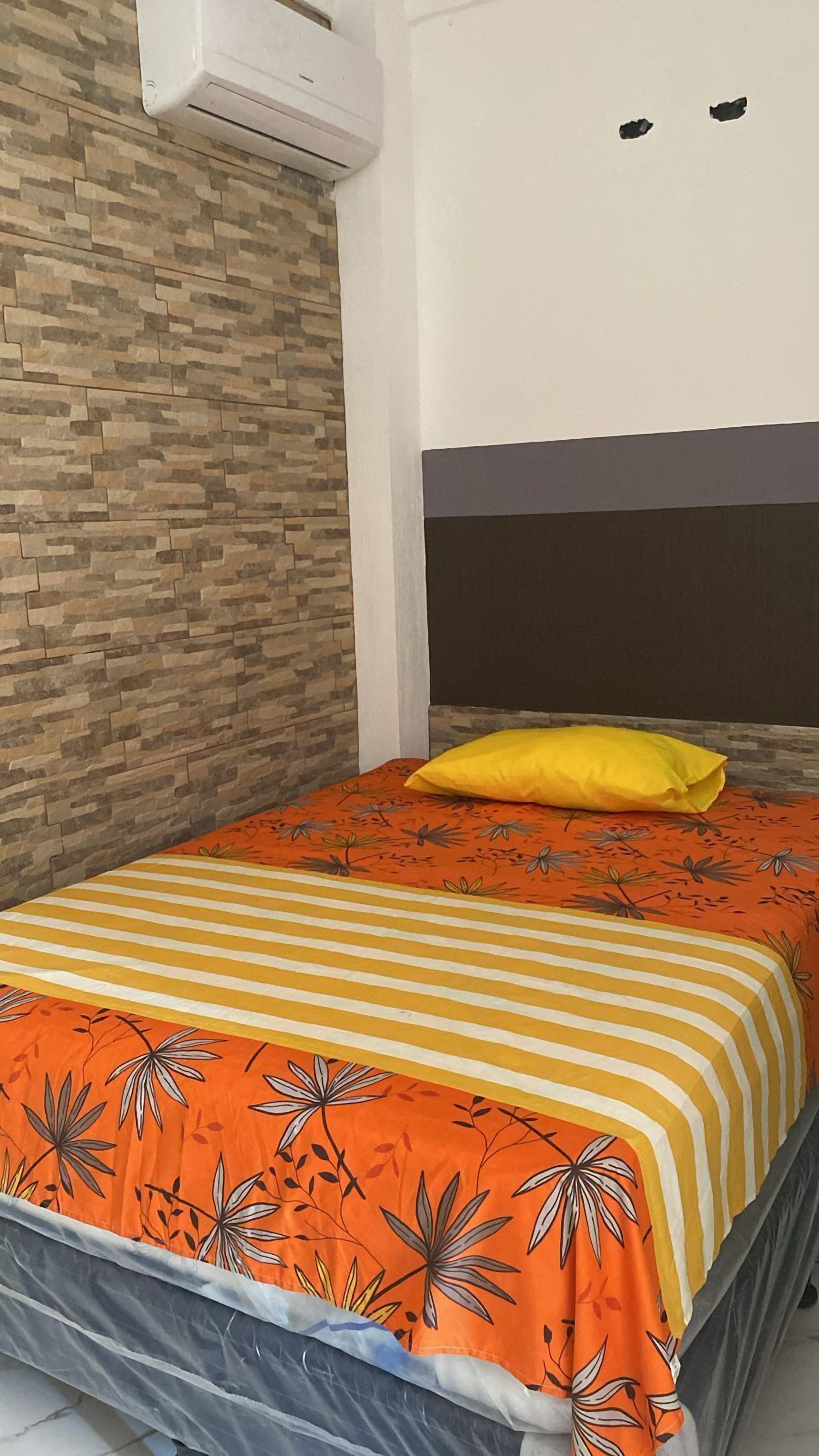 Quarto com cama, ar condicionado fixado na parede e colcha estampada.